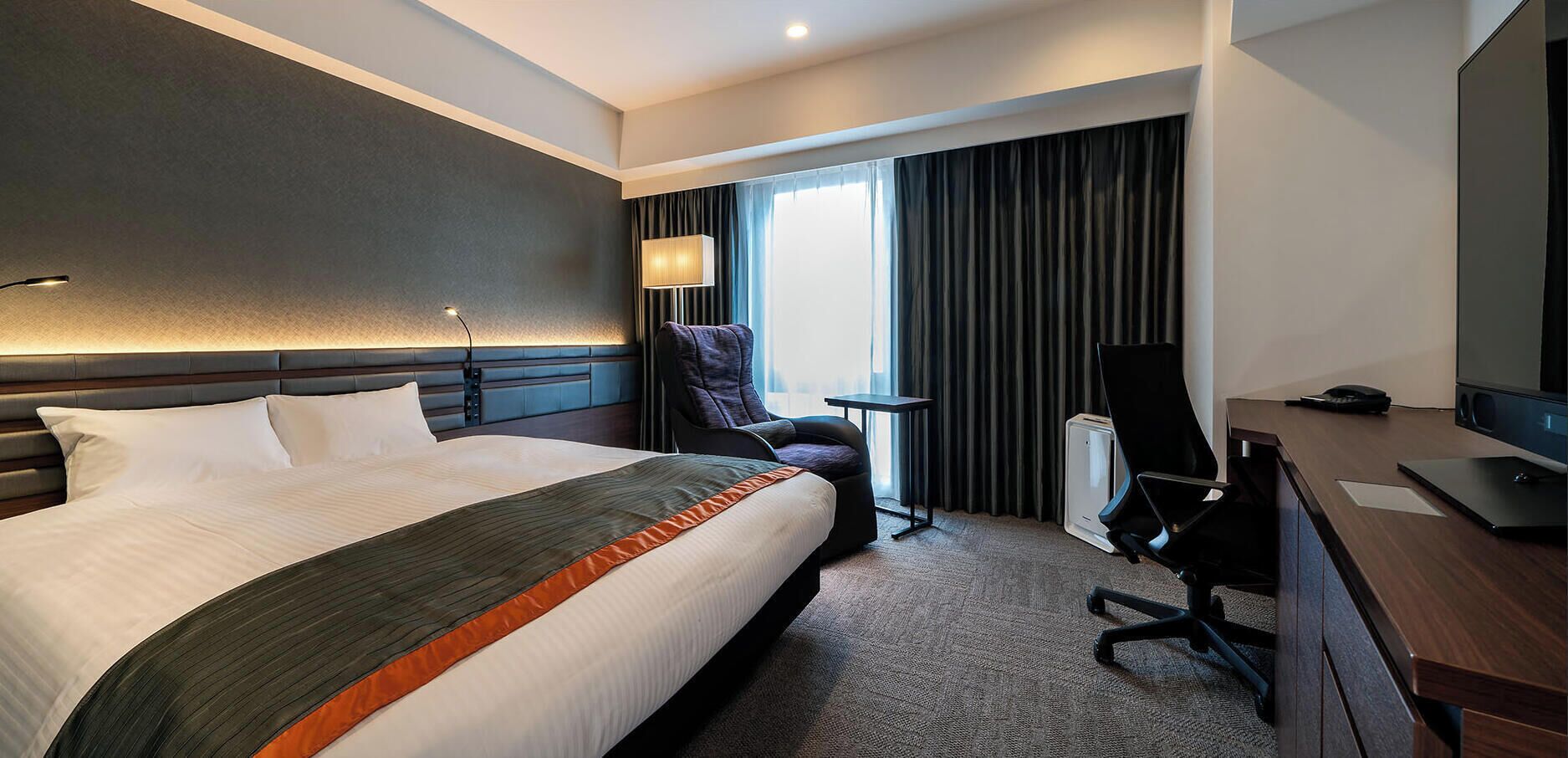 Quarto de hotel com uma cama grande, uma escrivaninha, uma cadeira, uma luminária e uma televisão.