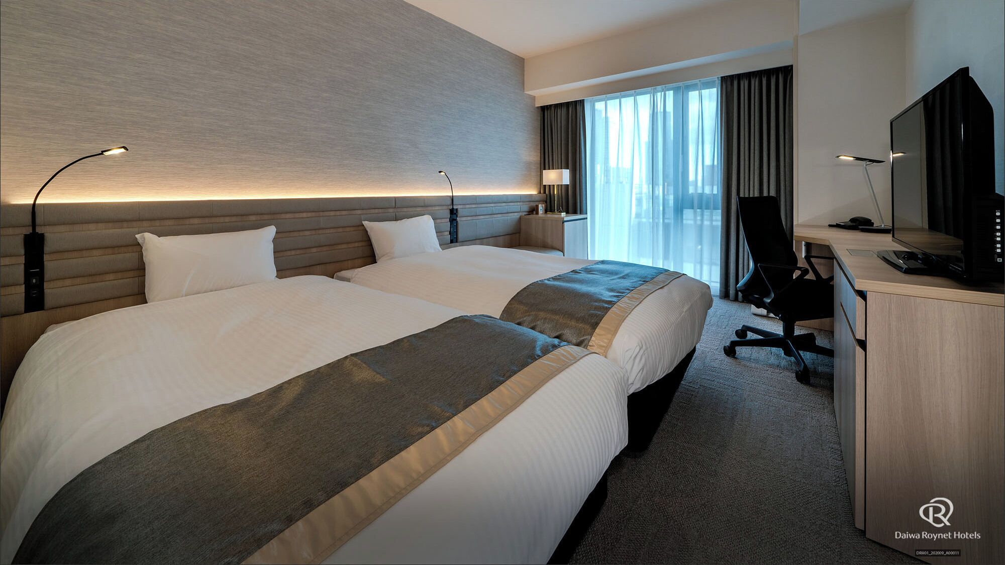 Quarto de hotel com duas camas, uma escrivaninha com computador e uma televisão.