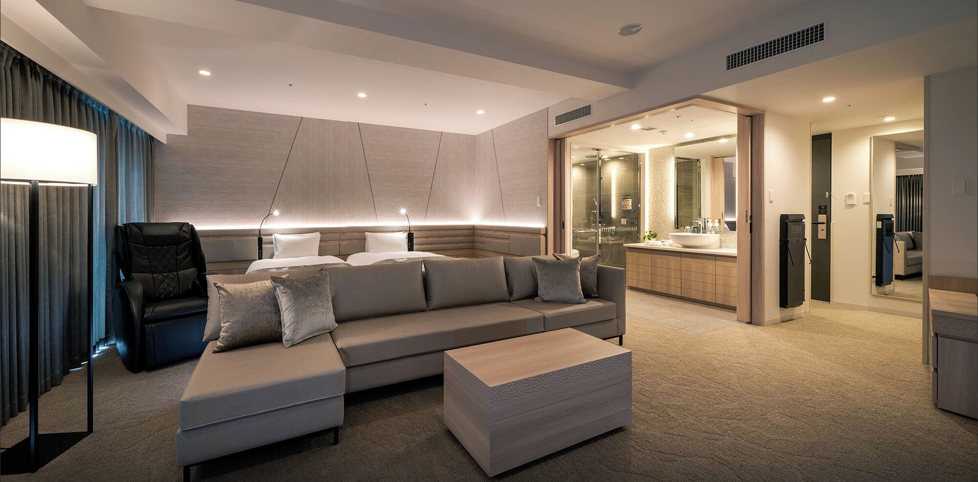 Quarto de hotel moderno com um amplo sofá em formato de L, uma poltrona individual, uma pequena mesa, um closet espaçoso, um banheiro com um espelho grande e área de chuveiro.