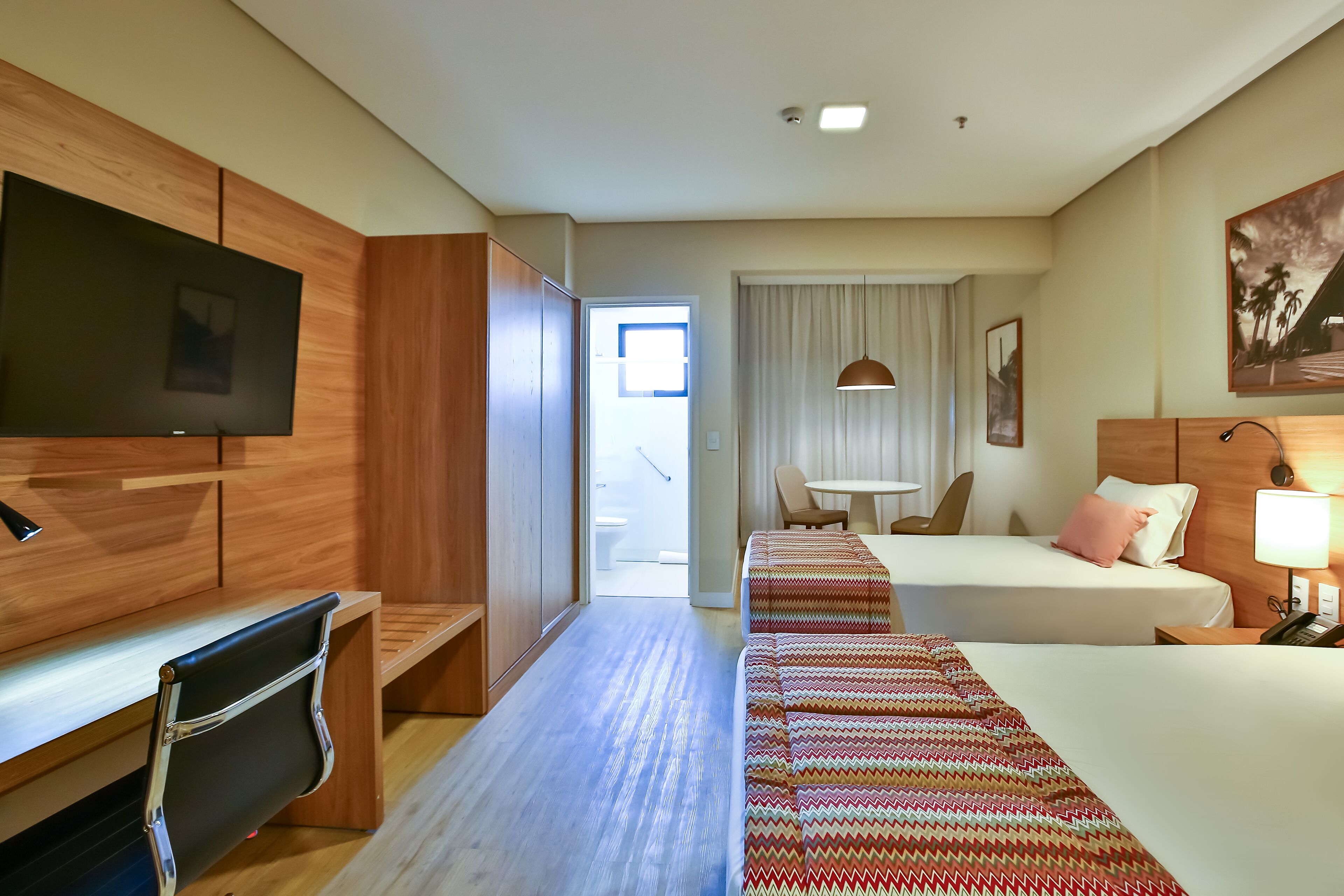 Quarto de hotel com televisão, mobiliário de madeira, escrivaninha, cama com roupa de cama estampada, mesa de cabeceira com abajur e um quadro na parede.