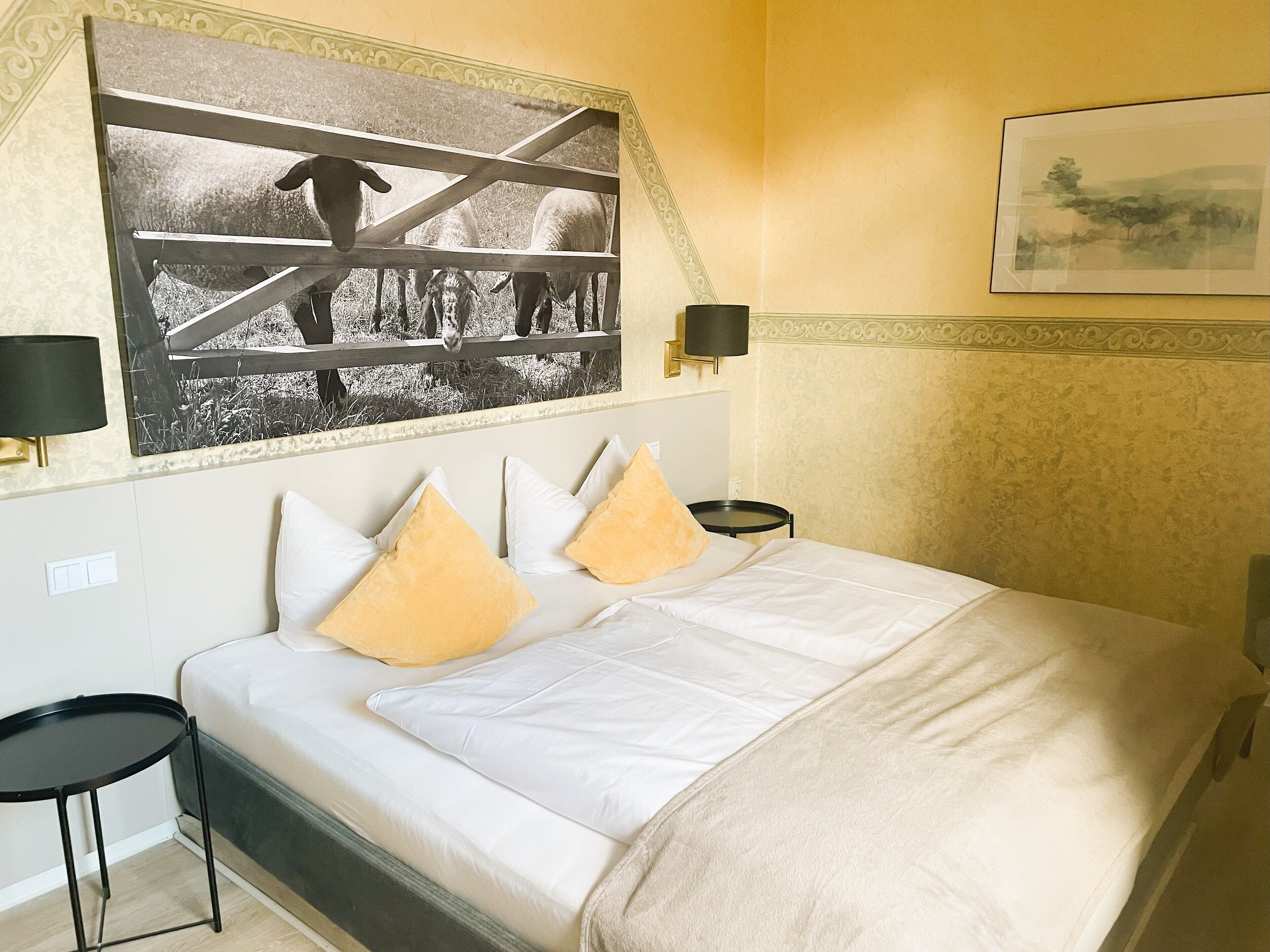Quarto de hotel com cama, uma pequena mesa, uma cadeira, um quadro na parede e um abajur.