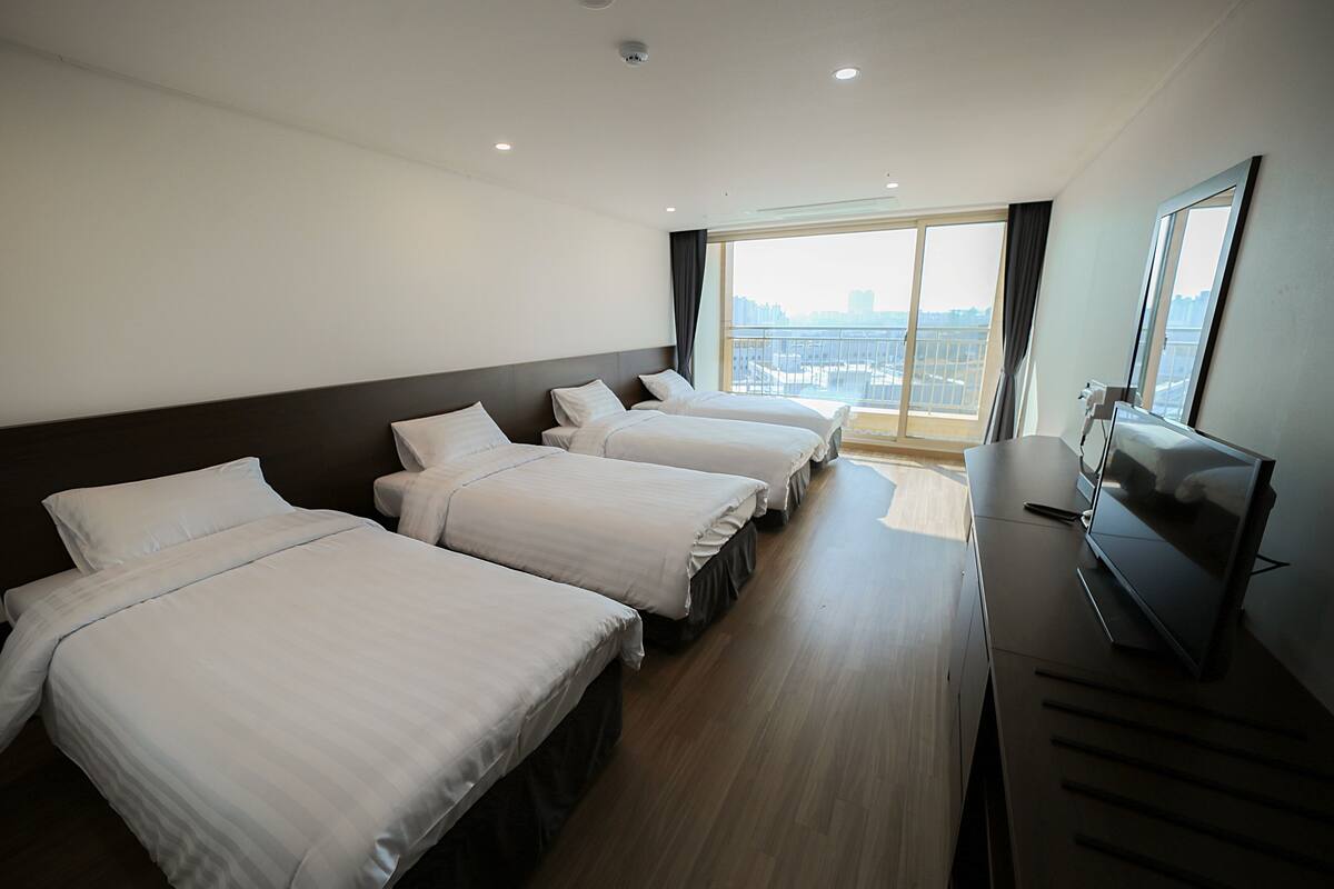 Quarto de hotel com três camas, uma televisão e uma janela grande com vista para a cidade.
