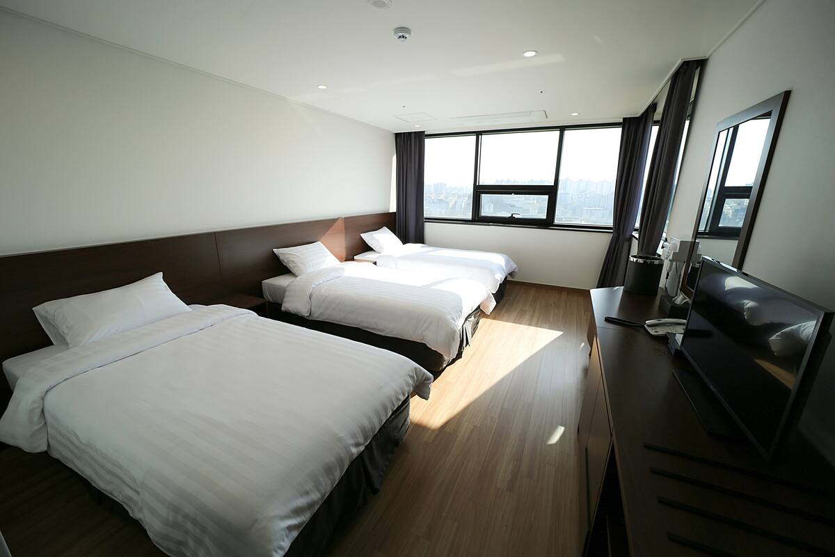 Quarto de hotel com três camas, uma mesa com computador e uma janela grande com vista para a cidade.