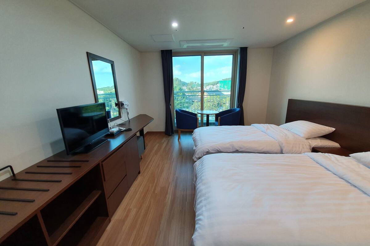 Quarto de hotel com uma cama, uma escrivaninha com televisão, uma cadeira e vista para o exterior.
