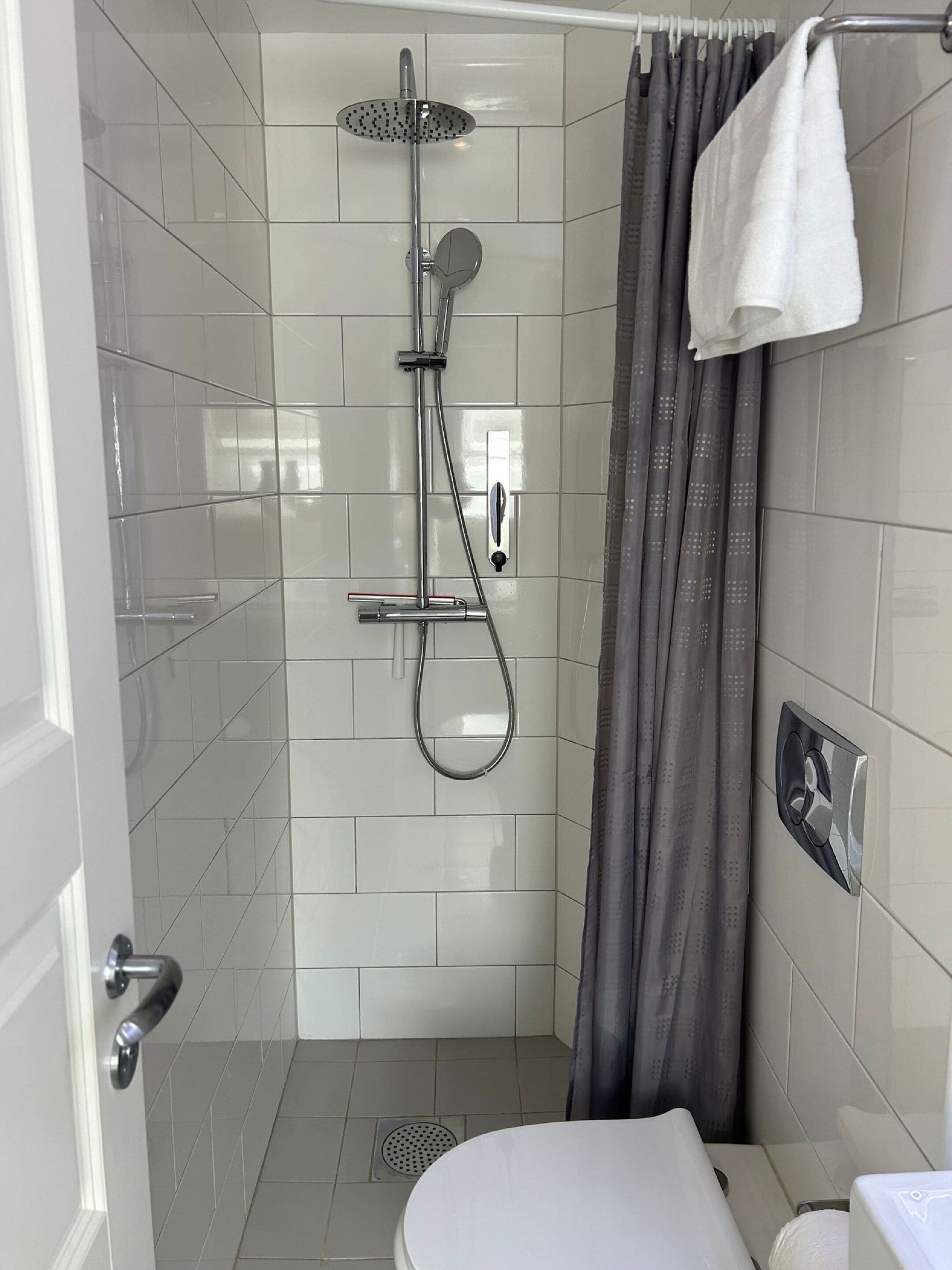 Um banheiro moderno com paredes revestidas de azulejos brancos, chuveiro com chuveiro manual, vaso sanitário e toalha pendurada em um porta-toalhas.