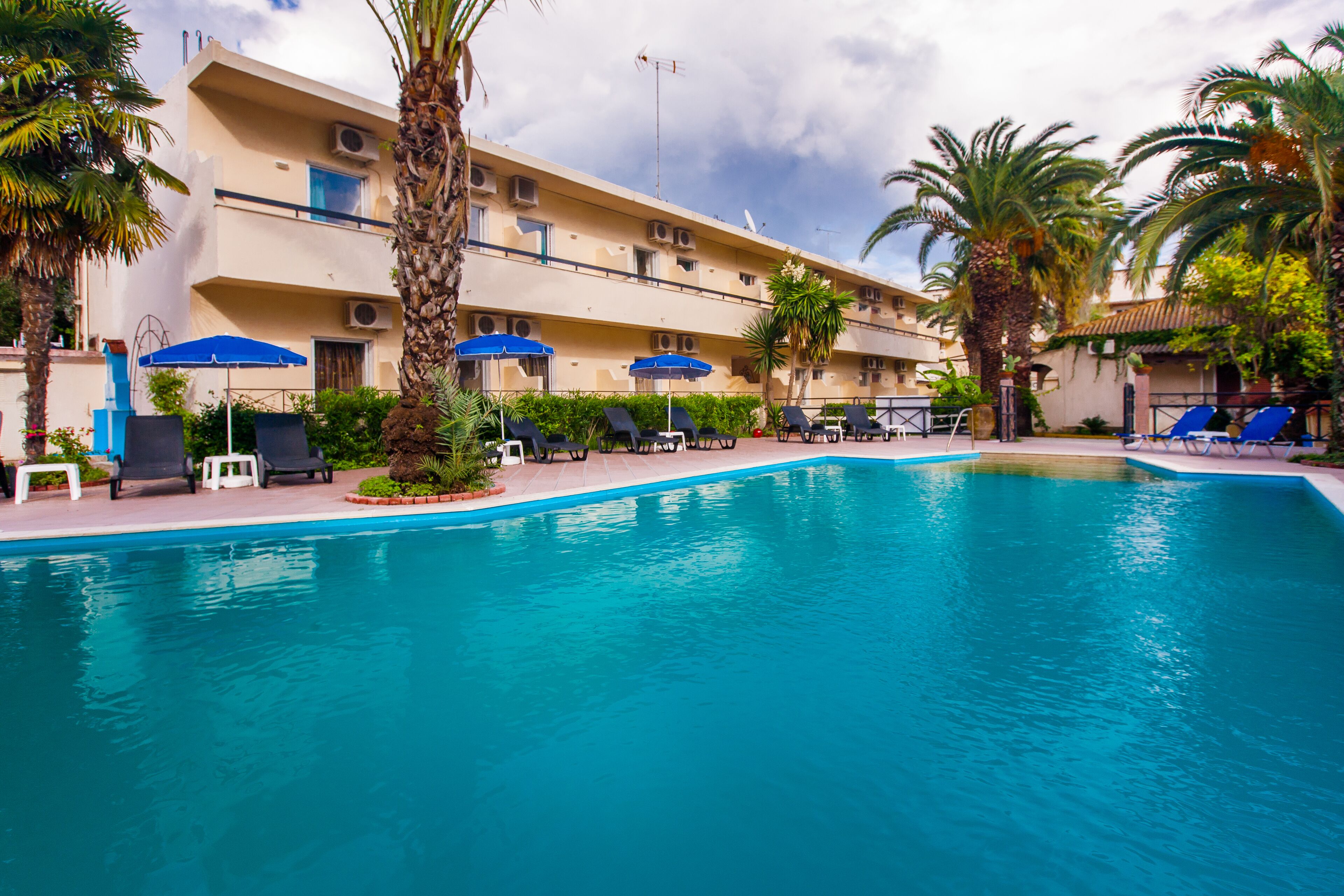 Piscina com espreguiçadeiras e guarda-sóis, cercada por palmeiras e um edifício com varandas.
