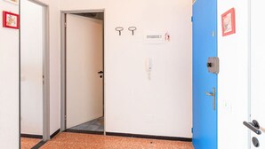 Um corredor com uma porta azul, uma porta branca e uma porta fechada. Duas chaves pendem na parede e há um termostato.