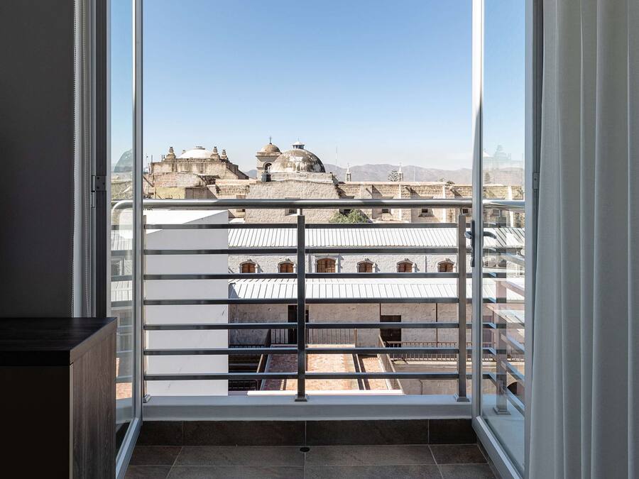Quarto com uma janela grande, varanda com guarda-corrimão de metal e vista para edifícios ao longe.