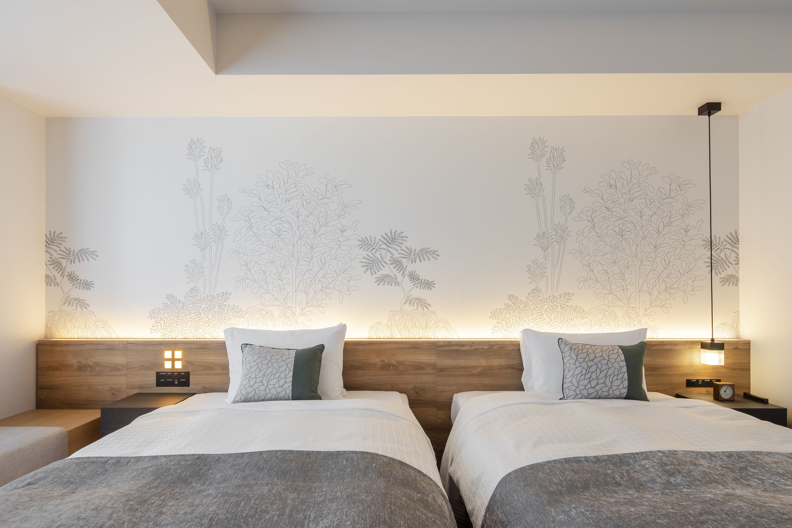 Quarto de hotel moderno com duas camas, uma cabeceira de madeira, um candeeiro fixado na parede e uma mesa de cabeceira com um relógio.