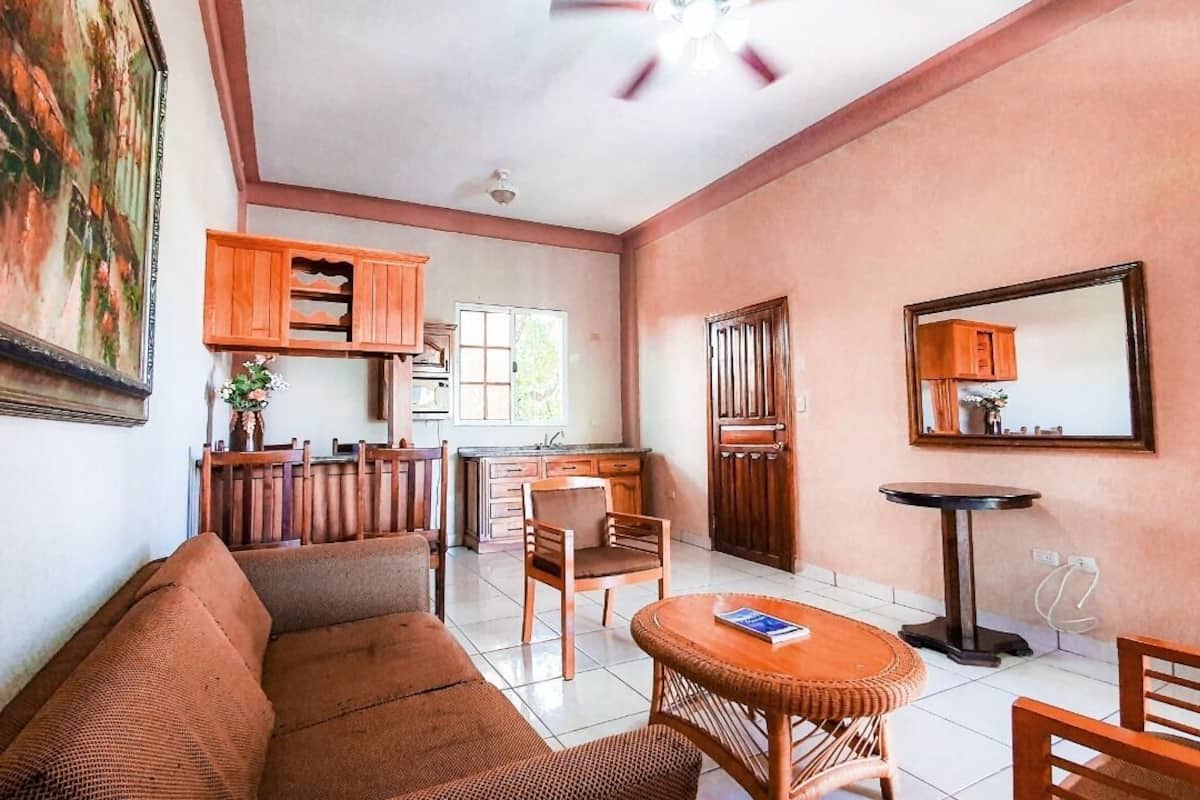 Uma sala de estar com sofá, cadeiras, uma mesinha redonda, armários de madeira, um quadro e uma janela.