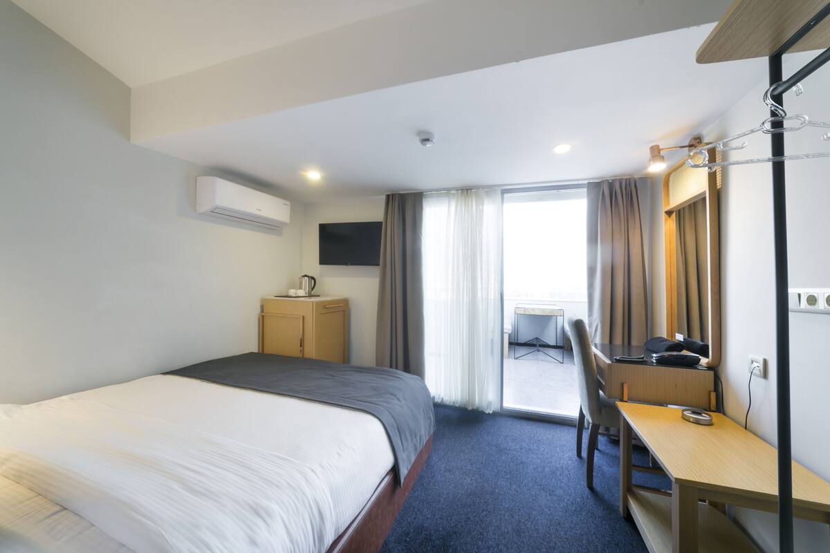 Quarto de hotel com cama, escrivaninha, cadeira, televisão e janela com cortinas.