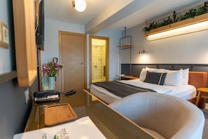 Quarto de hotel moderno com cama, mesa de cabeceira, cadeira e pequena escrivaninha com teclado e planta.