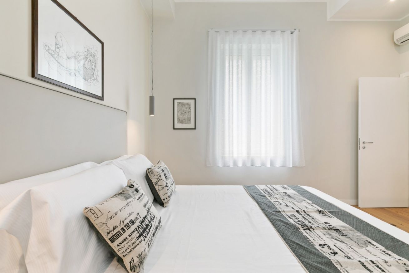 Quarto com cama, cortinas brancas, um quadro na parede e uma porta.