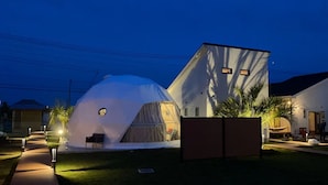 Uma tenda em formato de cúpula com uma pequena mesa e cadeira, um caminho que a leva até ela, e um edifício moderno com janelas amplas e telhado inclinado.