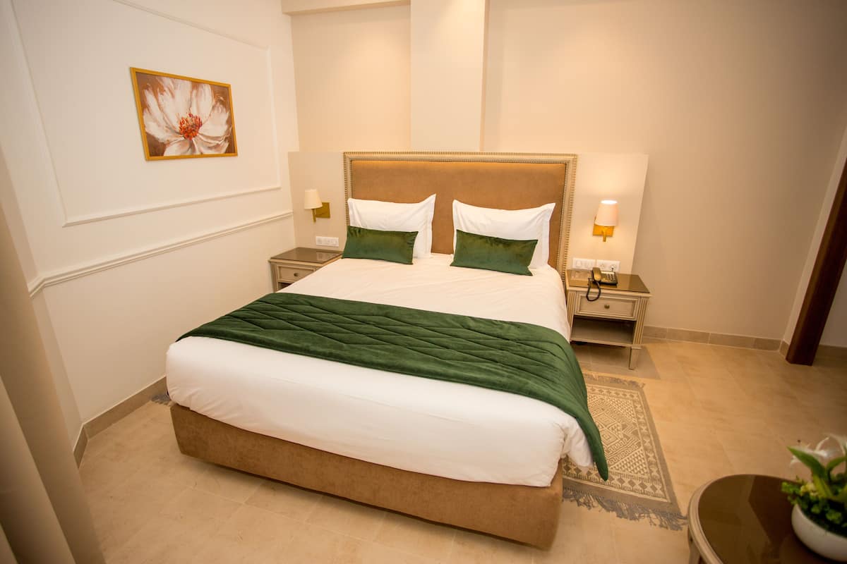 Quarto de hotel com uma cama grande, duas mesas de cabeceira, um criado-mudo, um cobertor verde e um quadro com flores na parede.