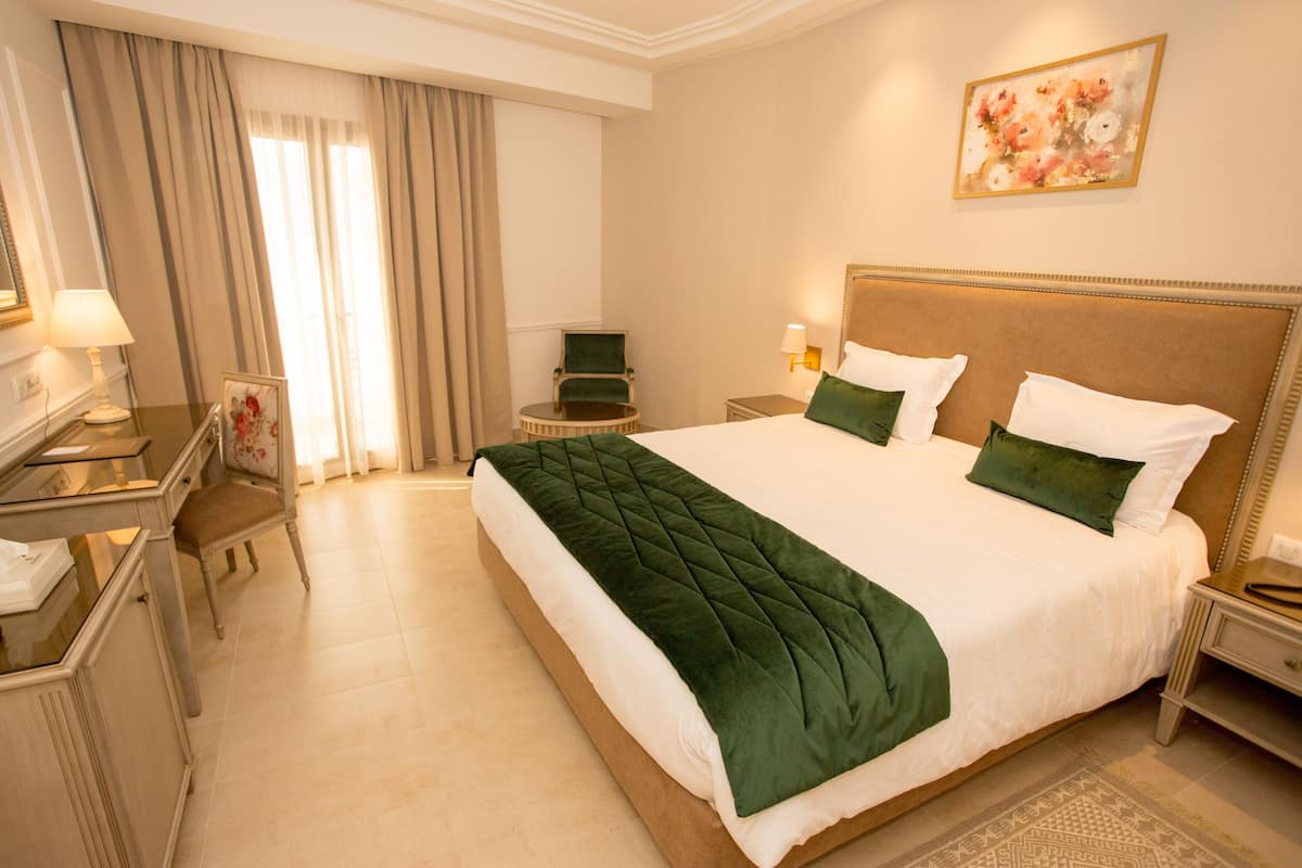 Quarto de hotel com uma cama grande, uma escrivaninha com luminária, uma cadeira, uma mesinha e um quadro na parede.