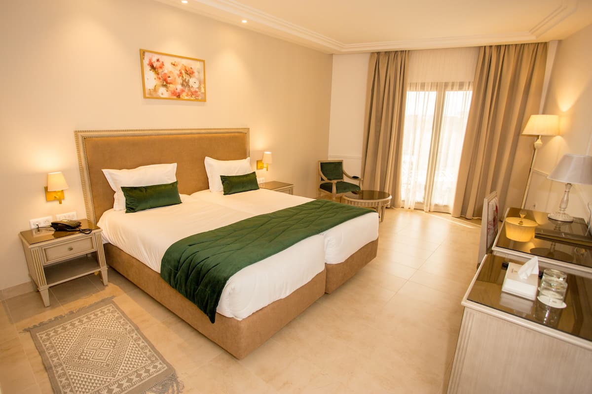 Quarto de hotel com uma cama grande, duas mesas de cabeceira com luminárias, uma cadeira, uma escrivaninha com luminária e uma janela com cortinas.
