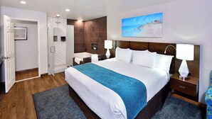 Quarto de hotel com uma cama grande, banheiro com chuveiro, mesa de cabeceira com luminária e um quadro na parede.