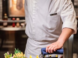 Um chefe de cozinha com uniforme branco e crachá, segurando um pano azul. Em primeiro plano, uma variedade de vegetais, incluindo milho, cenouras e brócolis.