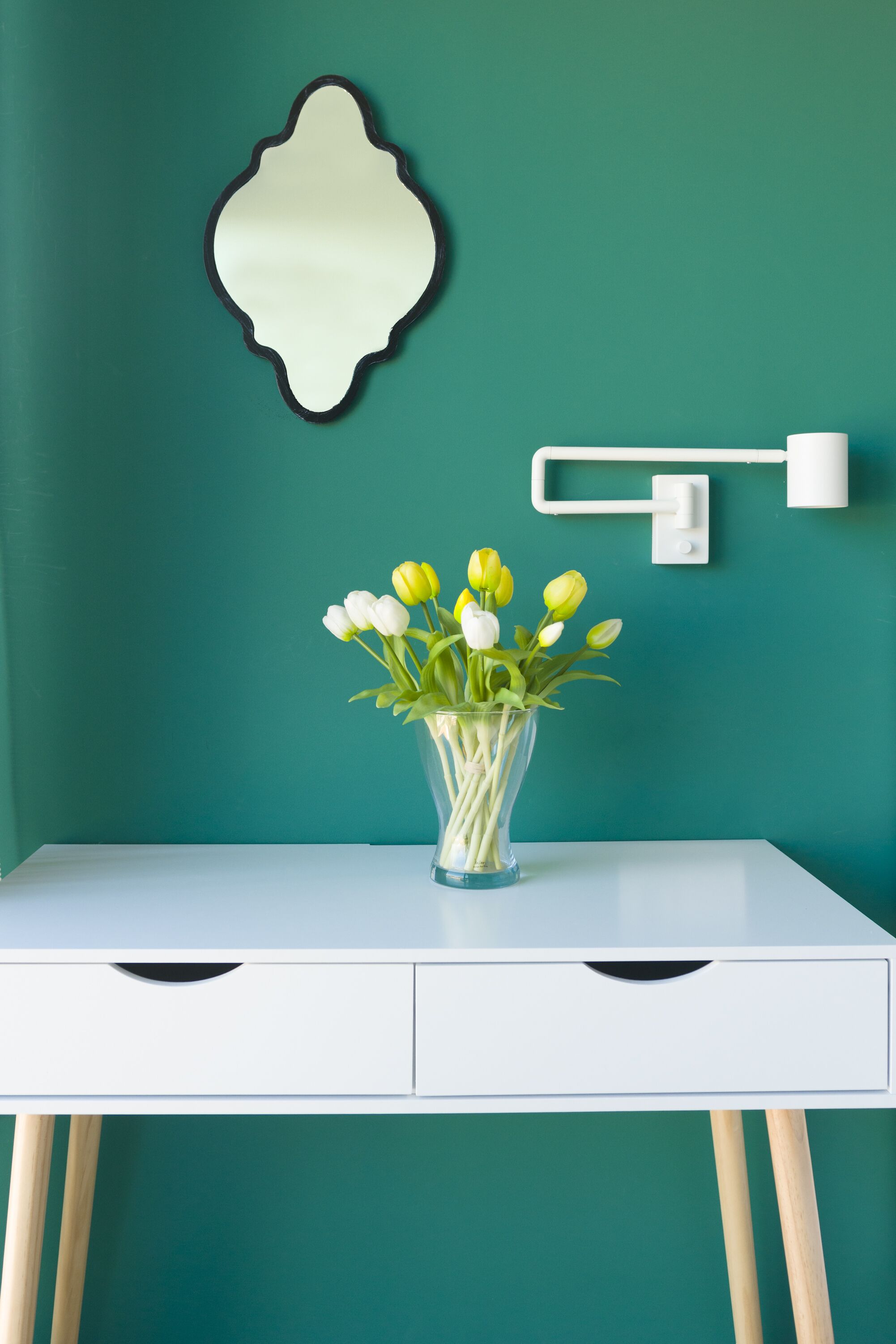 Uma secretária branca com um vaso de tulipas em frente a uma parede azul-turquesa, um espelho decorativo e um candeeiro fixado na parede.