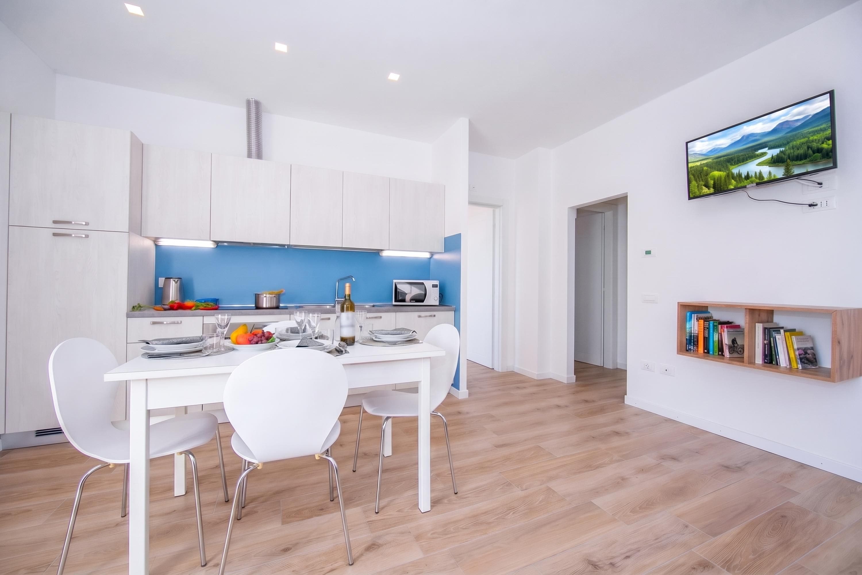 Cozinha moderna com armários brancos, painel de parede azul e área de refeições com mesa e cadeiras brancas.