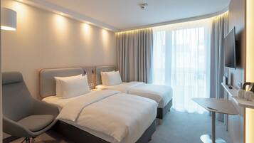 Quarto de hotel com duas camas, área de estar, televisão e janela com cortinas.