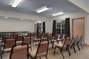 Sala de conferências com fileiras de cadeiras, uma tela de projetor e uma janela grande com cortinas.