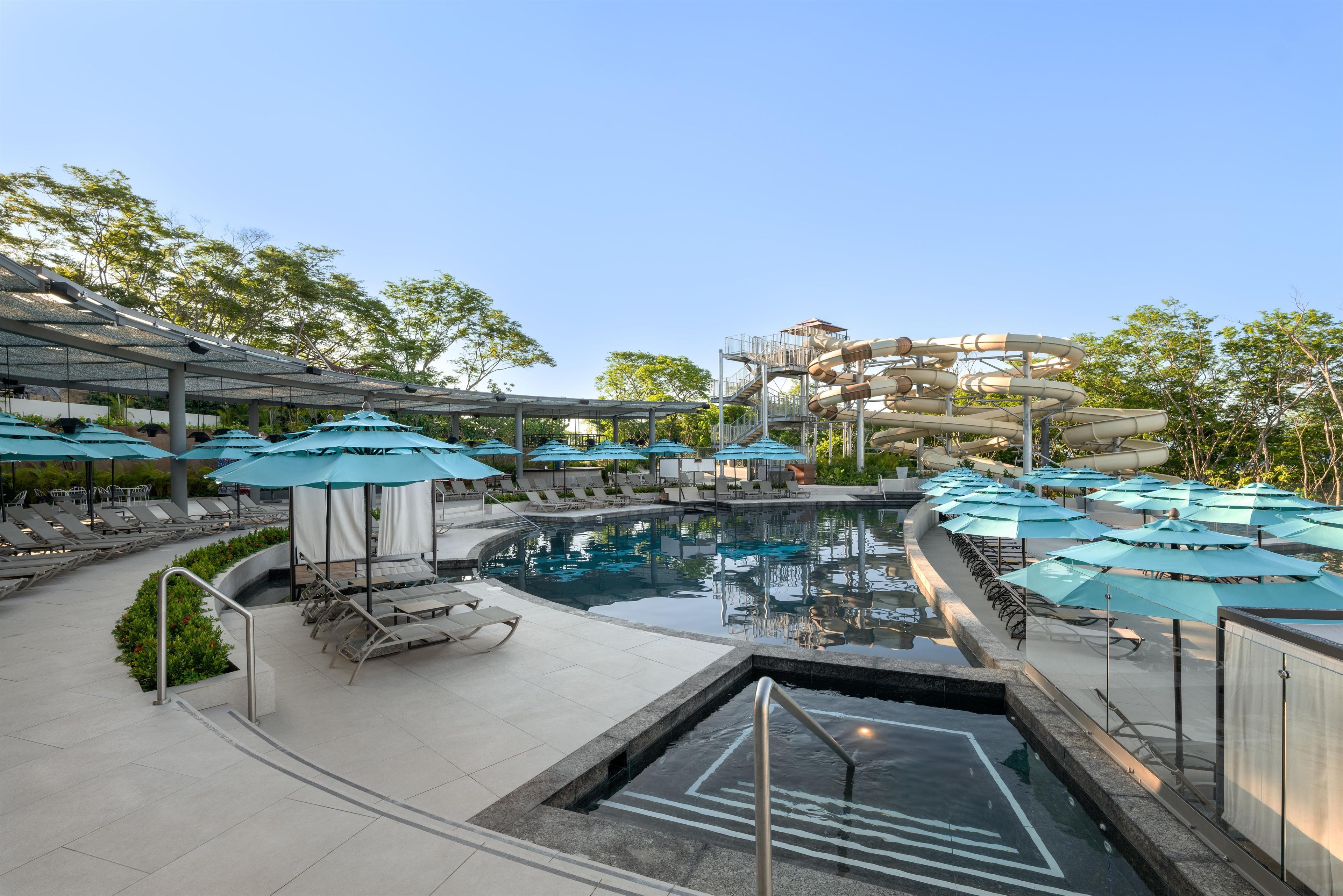 Área da piscina com espreguiçadeiras e guarda-sóis, um toboágua e um espaço coberto com assentos.