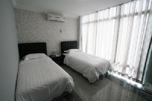 Quarto de hotel com duas camas, cabeceira escura, mesa de cabeceira pequena e janela com cortinas leves.