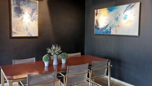 Uma sala de jantar com uma mesa de madeira e quatro cadeiras, três plantas em vasos sobre a mesa e duas pinturas abstratas nas paredes.