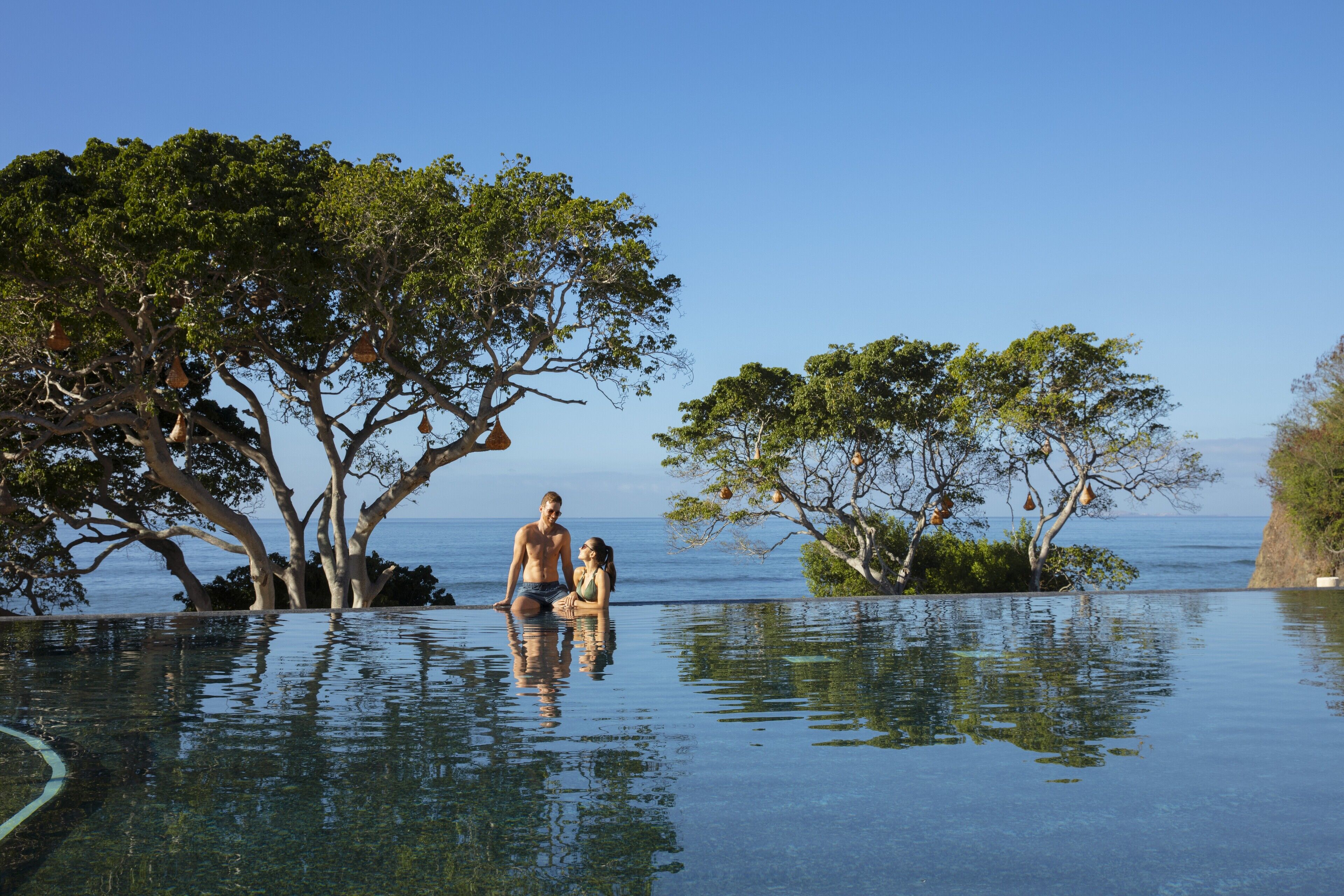 Um casal em uma piscina infinita transparente, com árvores e vista para o mar.