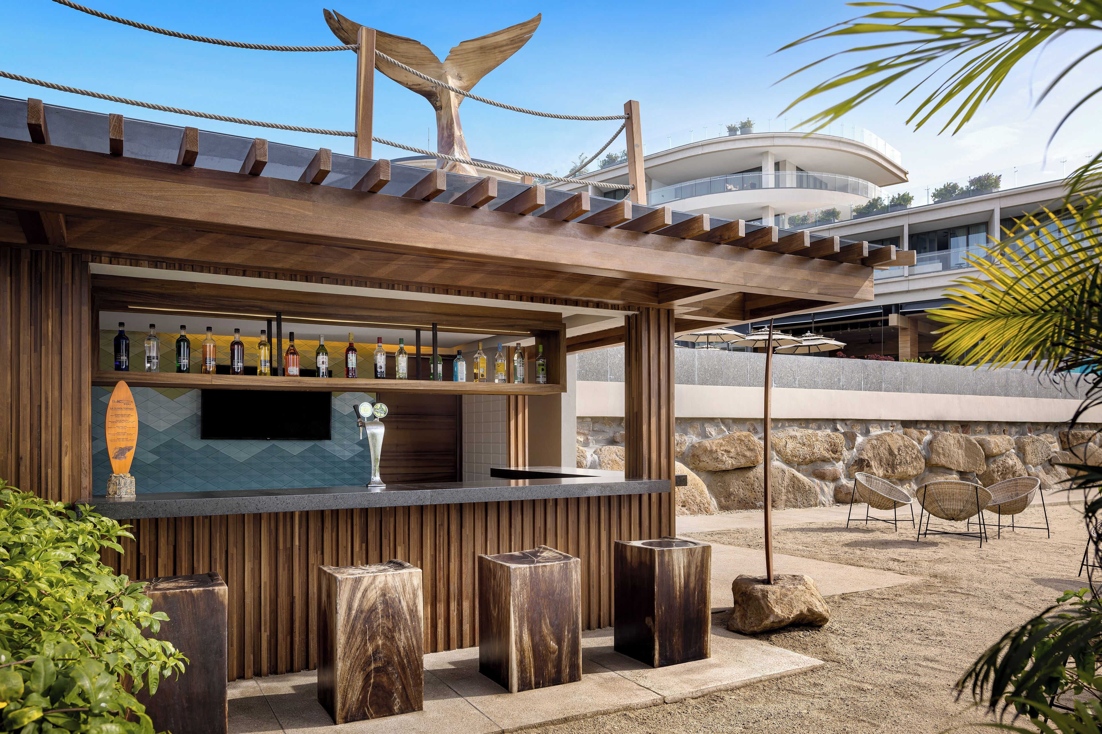 Um bar ao ar livre com ripas de madeira, uma escultura de baleia de madeira e uma parede de pedra ao fundo.