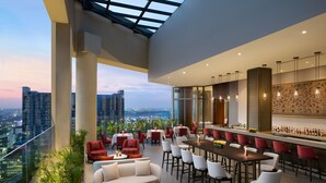 Um bar no terraço com mesas e cadeiras altas, balcão de bar e vista para a cidade.