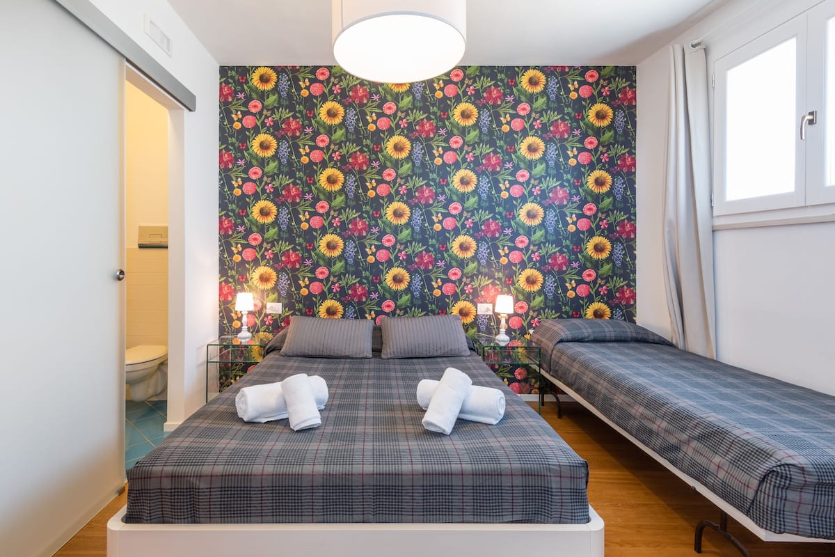 Quarto de hotel com cama, duas mesas de cabeceira, uma mesinha de apoio, uma janela e papel de parede floral.