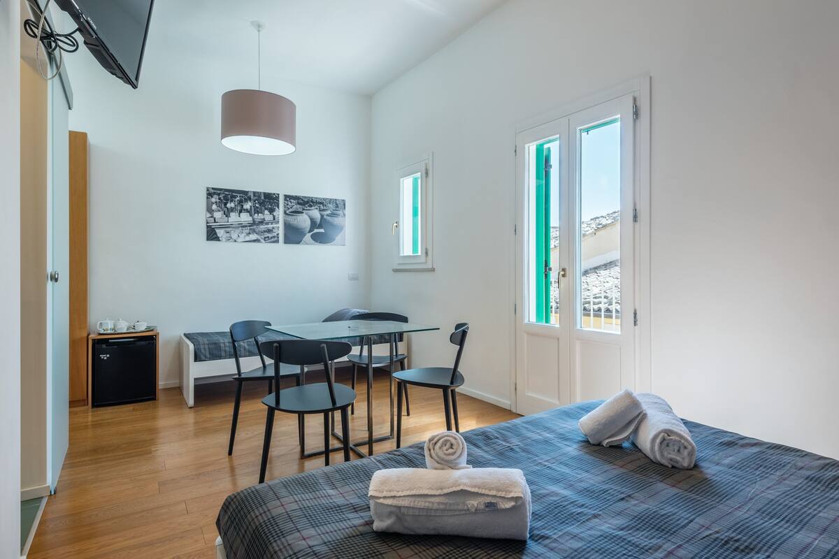 Quarto moderno com cama, mesinha, cadeiras, escrivaninha e geladeira.