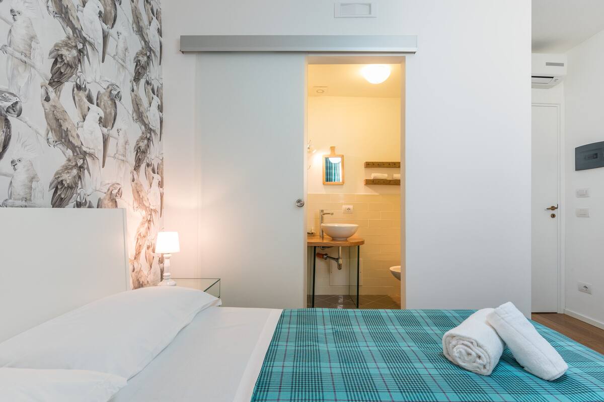 Quarto de hotel com cama, banheiro com pia e espelho, e parede com mural de aves.
