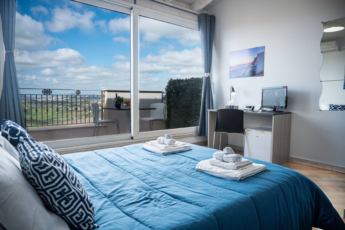 Quarto de hotel com uma cama grande, uma escrivaninha com computador e vista para o campo através de uma varanda.