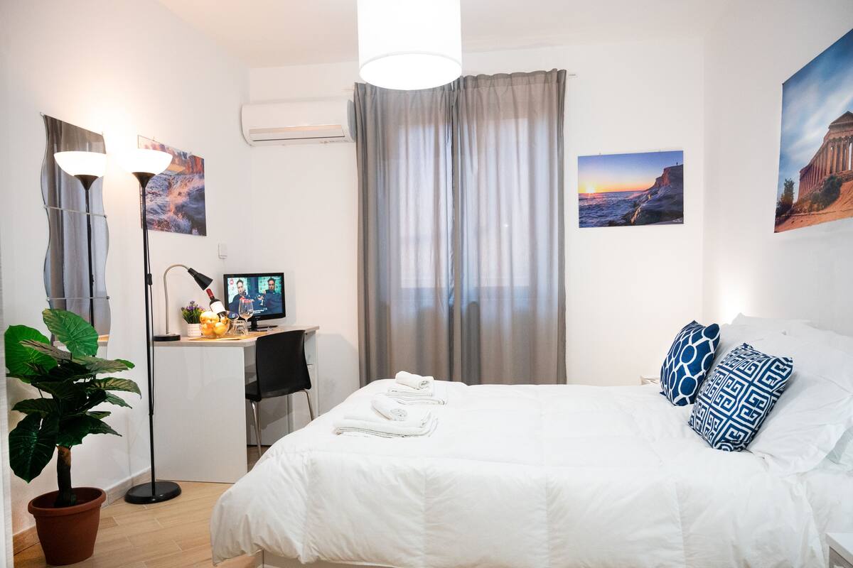 Quarto de hotel com cama, escrivaninha, computador, planta em vaso e obras de arte nas paredes.