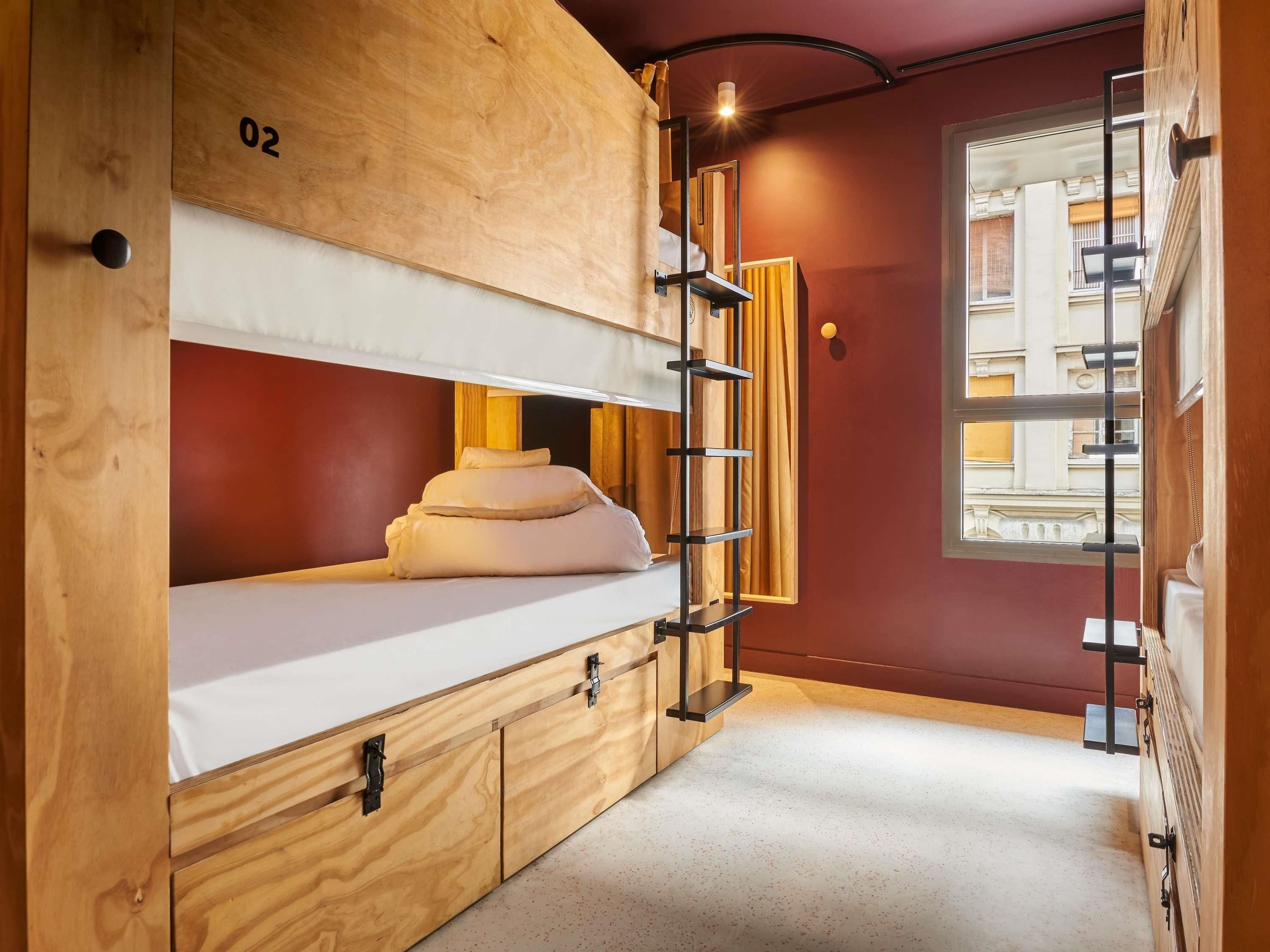 Quarto compacto com beliche, paredes de madeira, janela com cortinas e vista para prédios.