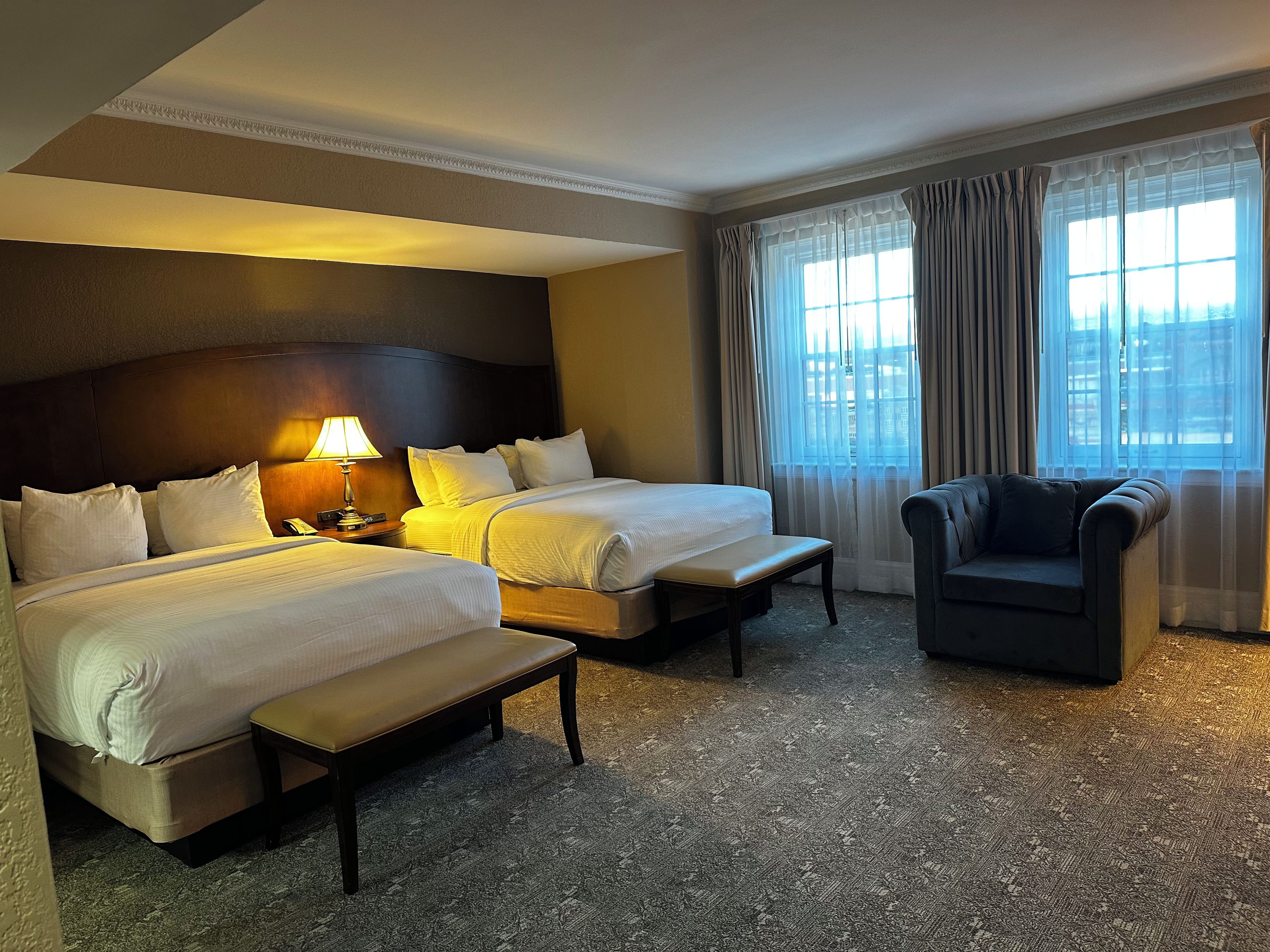 Quarto de hotel com duas camas, uma mesa, uma cadeira e uma janela grande com cortinas.
