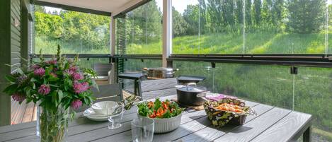 Mesa de jantar posta com comida e bebidas em um deck com vista para um campo gramado e árvores.