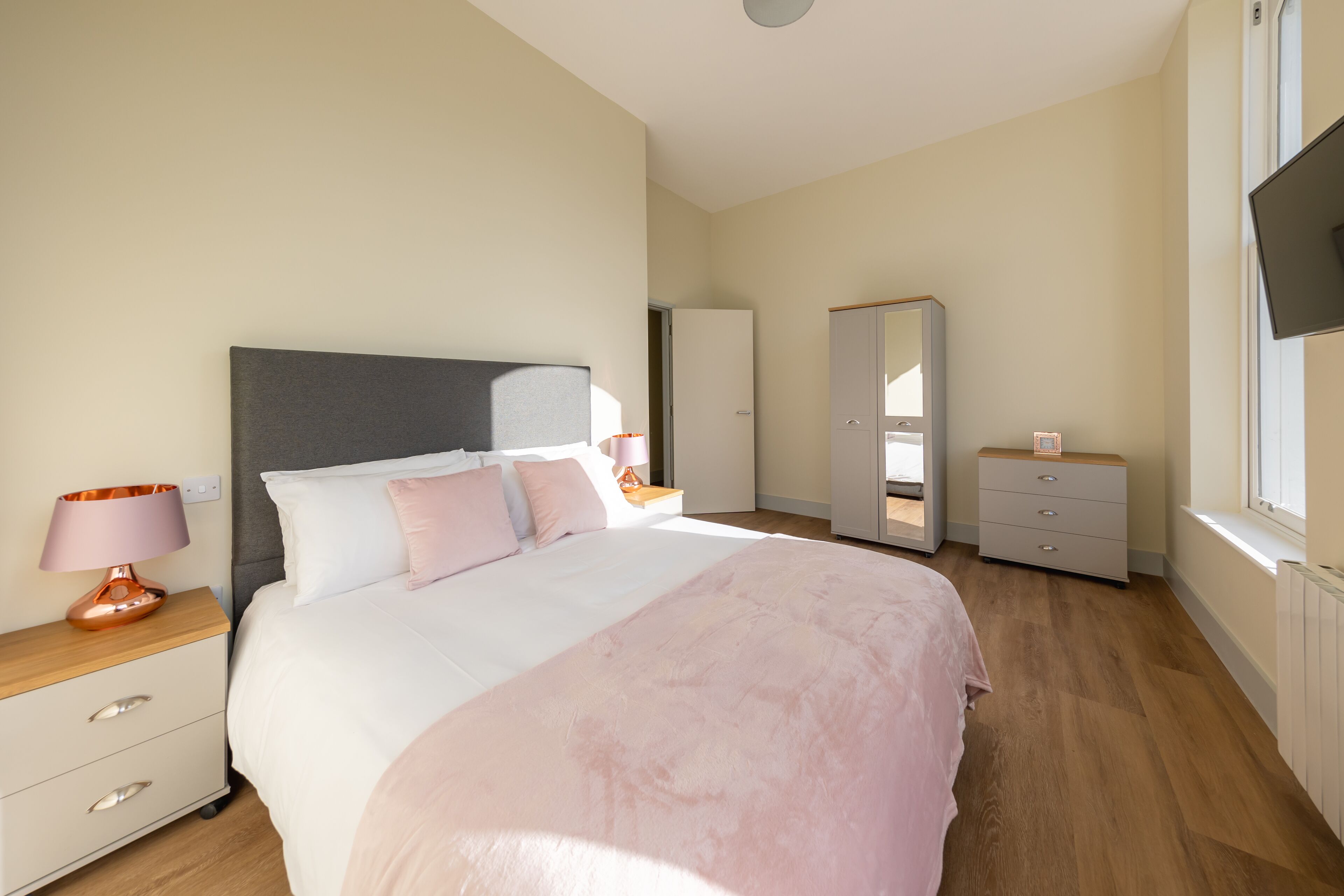 Quarto com cama, mesas de cabeceira, cômoda, guarda-roupa e televisão fixada na parede.
