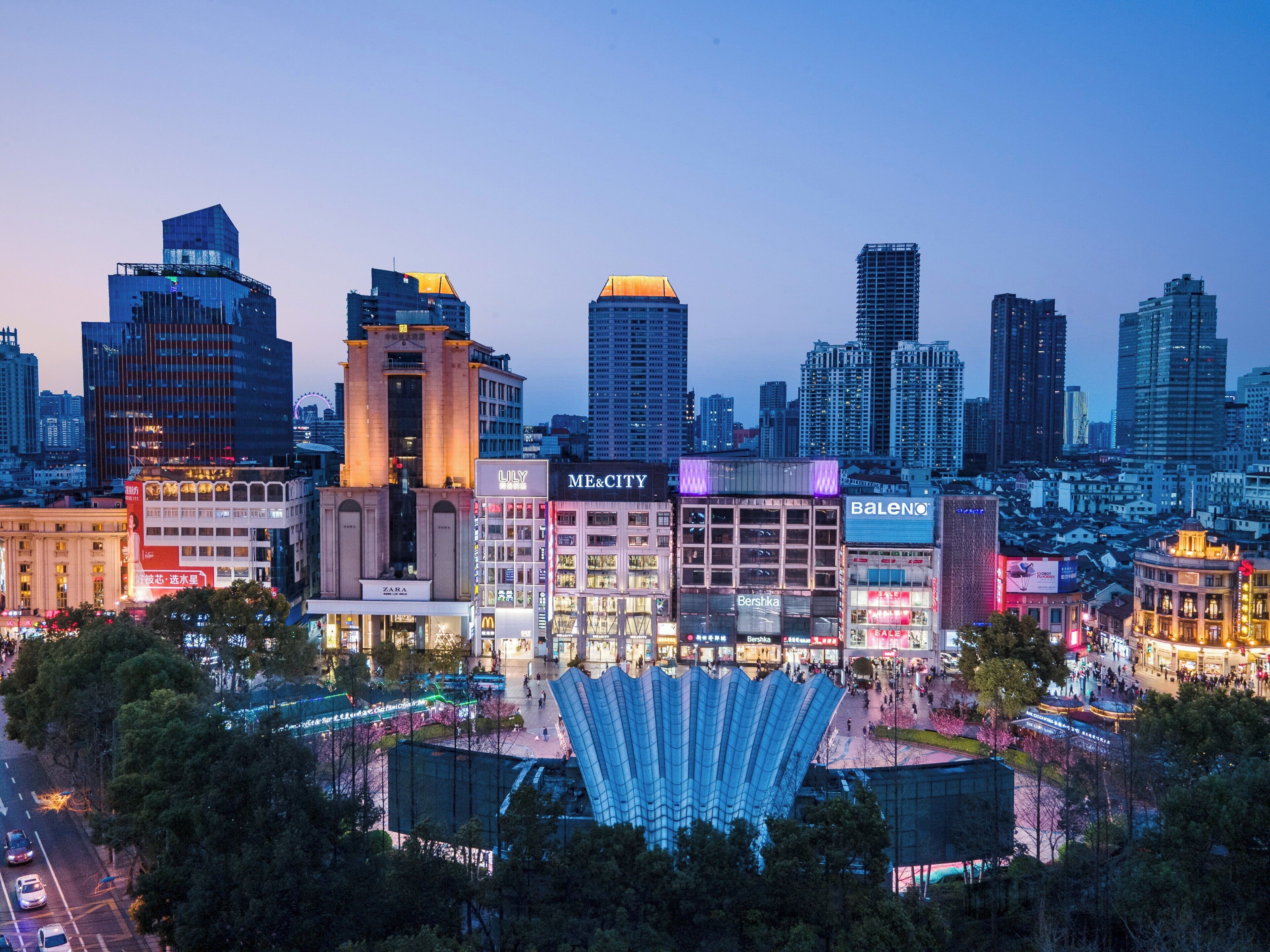 Paisagem urbana com edifícios altos e modernos, um centro comercial com nomes de lojas visíveis e uma estrutura azul marcante em primeiro plano.
