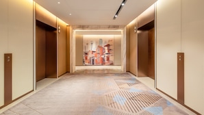 Corredor com elevadores, um tapete estampado e uma obra de arte com paisagem urbana na parede.
