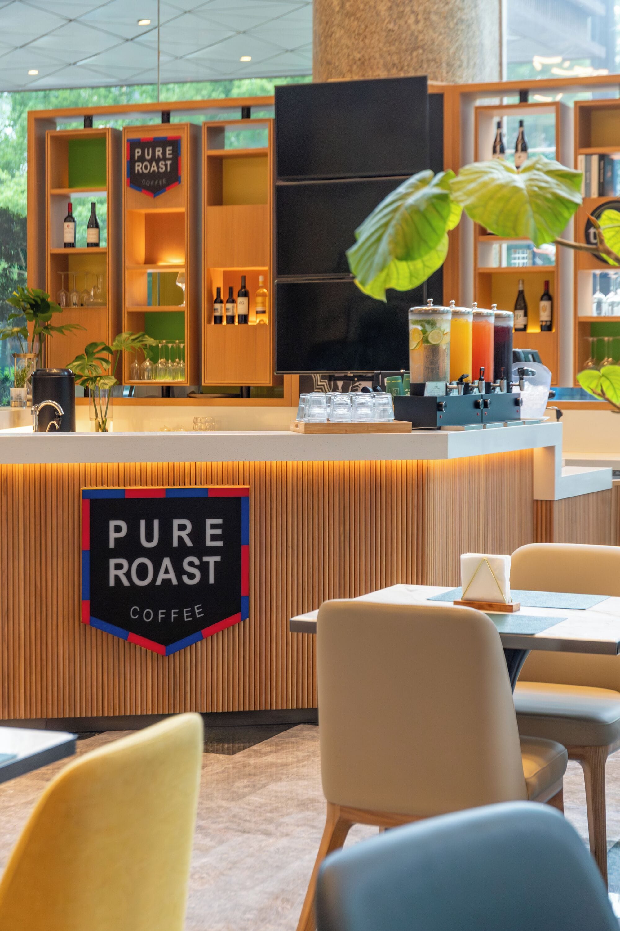 Balcão de café com um letreiro que diz 'PURE ROAST COFFEE', prateleiras com garrafas, uma televisão e área de estar.