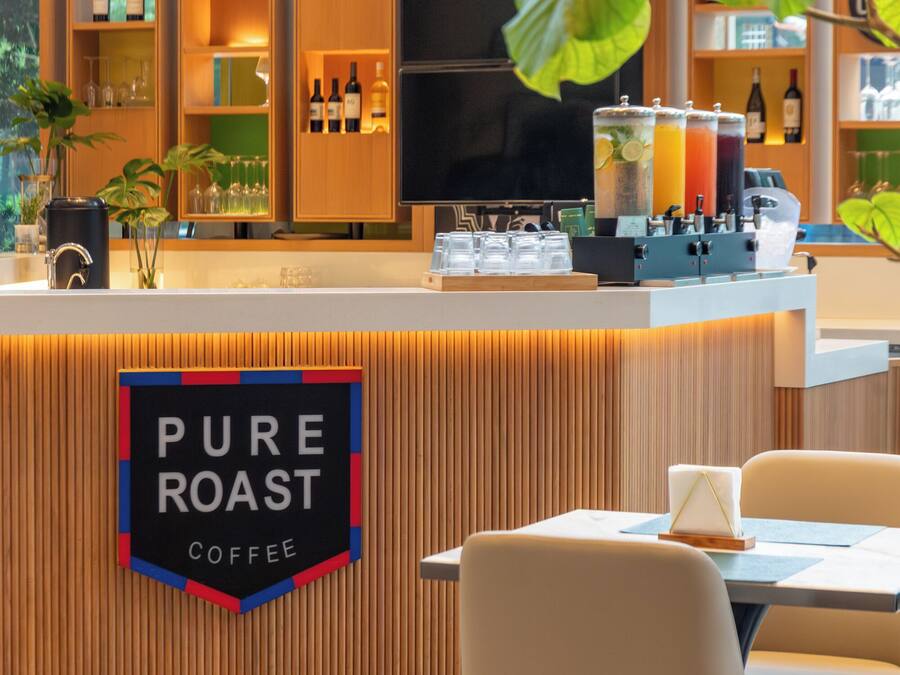 Balcão de café com um letreiro que diz 'PURE ROAST COFFEE', prateleiras com garrafas, uma televisão e área de estar.