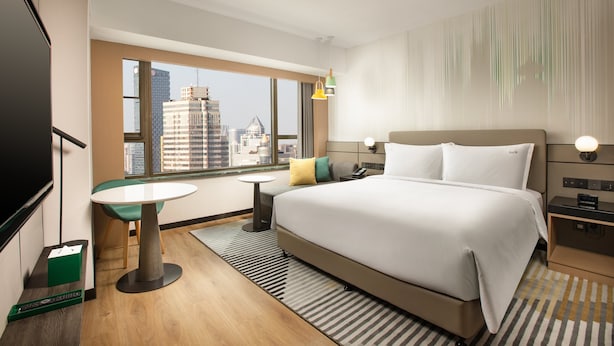 Quarto de hotel com uma cama grande, uma mesa pequena, uma cadeira, uma televisão e uma janela com vista para a cidade.