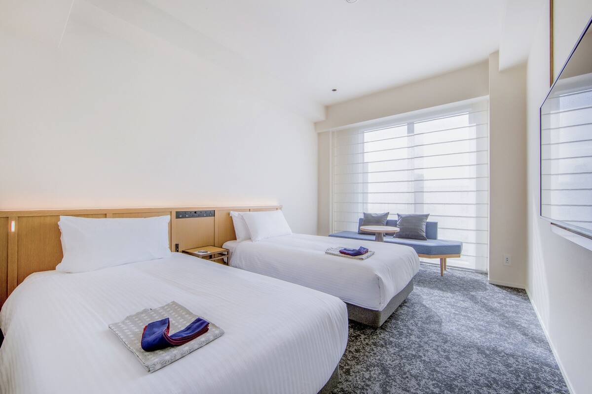 Quarto de hotel com duas camas, uma mesinha e uma janela com persianas.