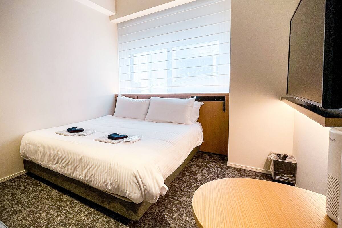 Quarto de hotel com cama, mesa pequena, televisão e janela com persianas.