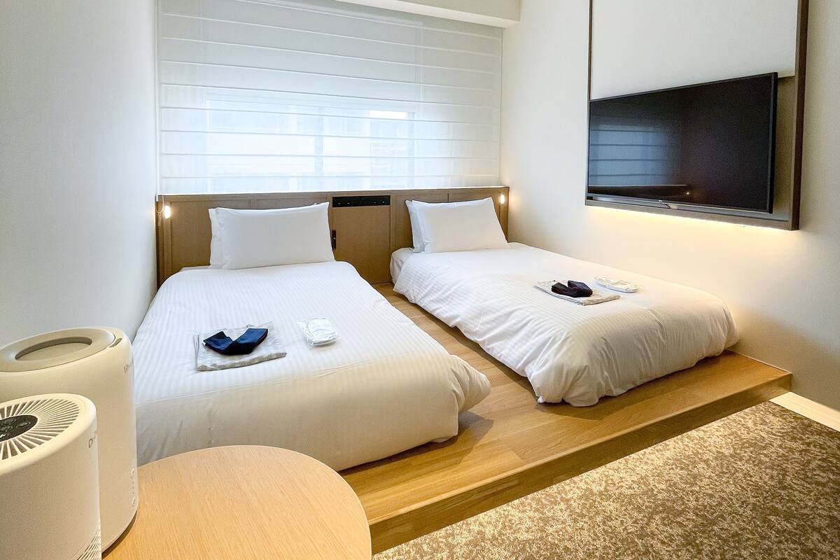 Quarto de hotel com duas camas, televisão, mesa de cabeceira e janela com persianas.